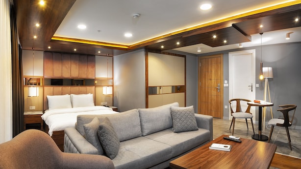 Quarto de hotel moderno com cama, sofá, poltrona, teto de madeira e uma pequena mesa com um controle remoto.