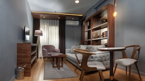 Uma sala de estar moderna com um sofá cinzento, mesa de centro de madeira e cadeiras a combinar. Há uma estante com objetos decorativos e uma TV fixada na parede.