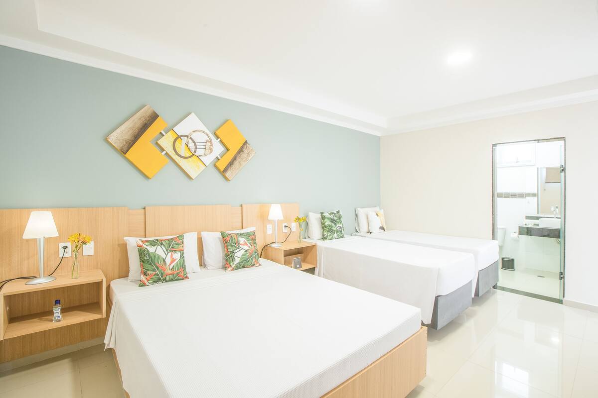 Quarto de hotel com duas camas, uma cabeceira de madeira, mesa de cabeceira com luminária e uma obra de arte na parede.