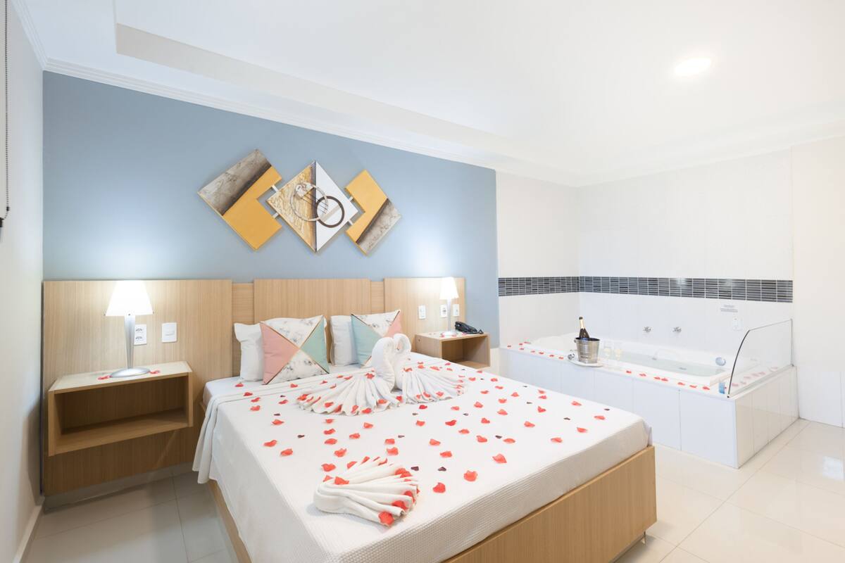 Quarto de hotel moderno com uma cama grande, mesa de cabeceira, quadro na parede, banheiro com banheira e porta-toalhas.