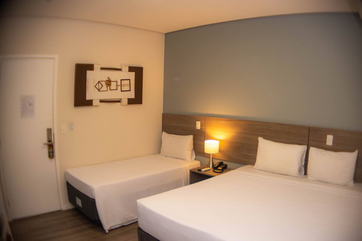 Quarto de hotel com duas camas de solteiro, uma mesa de cabeceira com abajur e uma obra de arte pendurada na parede.