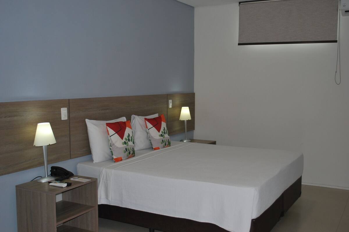 Cama bem arrumada com roupa de cama branca, duas luminárias de cabeceira e um painel de madeira.