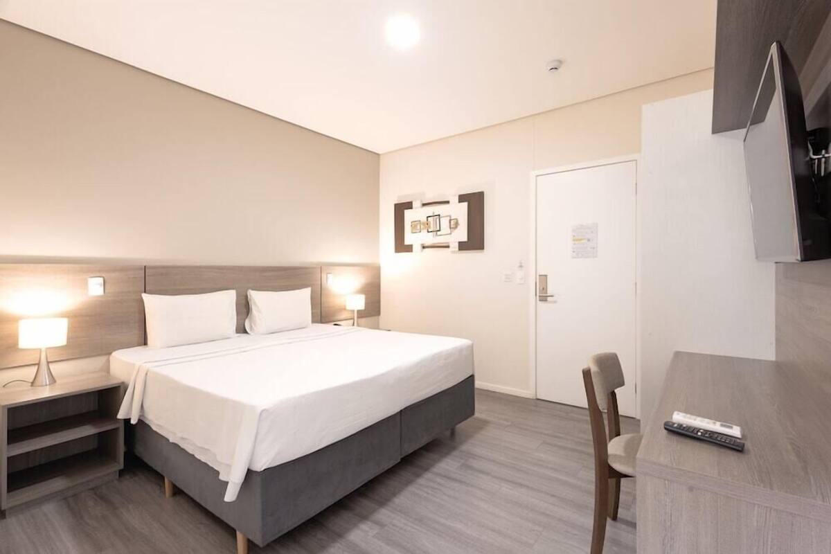 Quarto de hotel com cama, luminárias de cabeceira, escrivaninha e televisão.