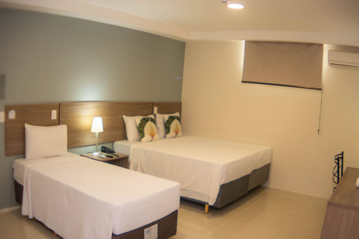 Quarto de hotel com duas camas de solteiro, uma mesa de cabeceira com luminária, uma pequena mesa e um ar condicionado fixado na parede.