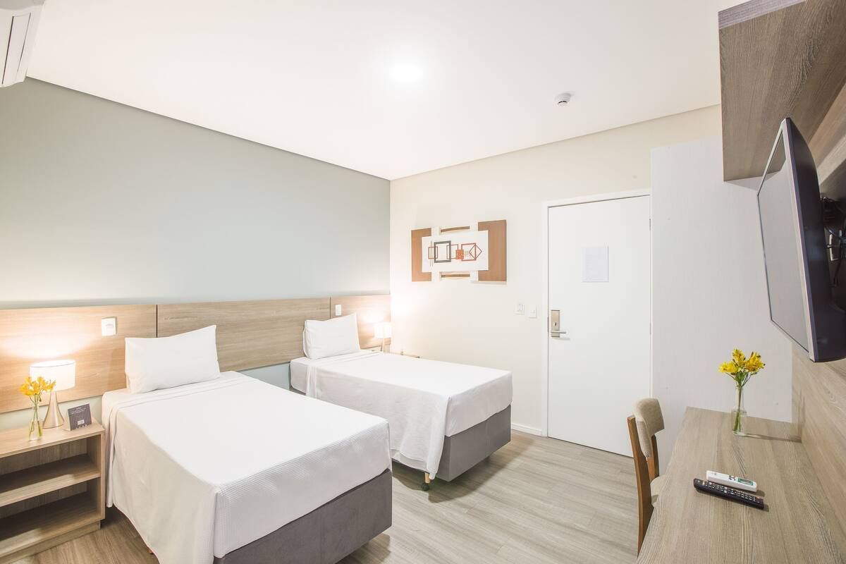 Quarto de hotel com duas camas, uma escrivaninha, uma cadeira, uma televisão e uma porta.