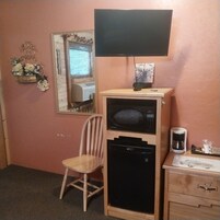 Quarto compacto com cadeira de madeira, pequena geladeira preta, máquina de café e televisão de tela plana fixada na parede.
