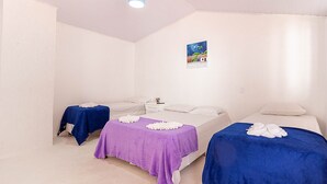 Quarto com três camas, cada uma com um espalha-cama de cor diferente. Há uma pequena mesa com abajur e um quadro na parede.
