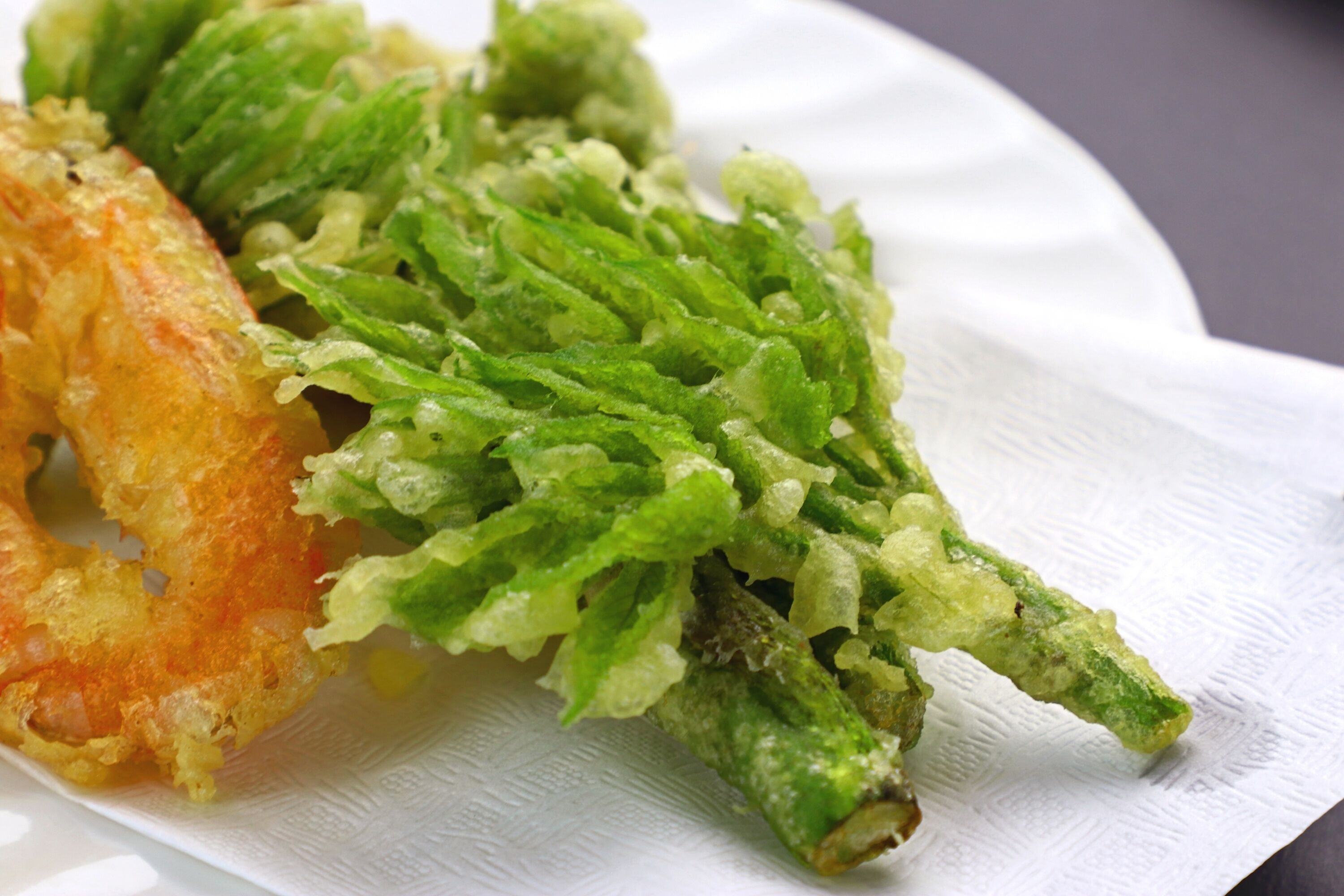 Prato com legumes fritos, incluindo um pedaço de couve-flor e um legume verde, possivelmente quiabo, sobre um guardanapo de papel branco.