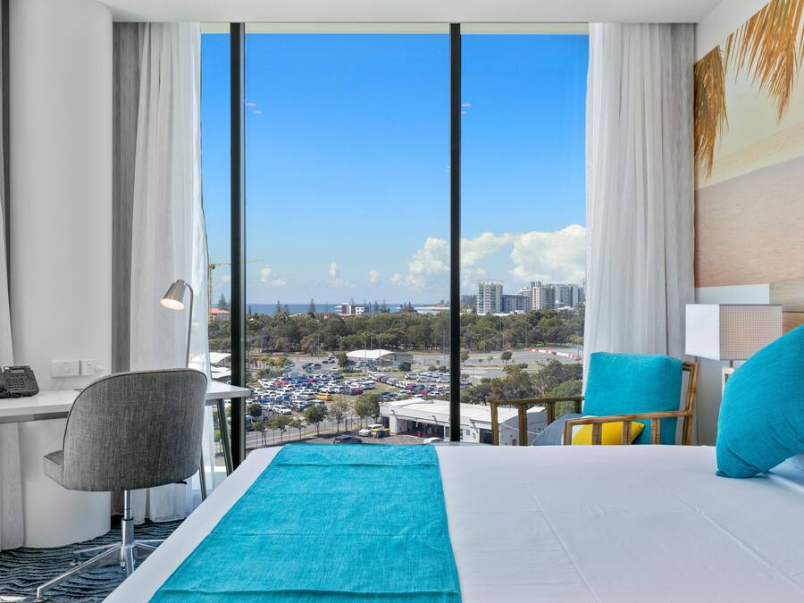 Quarto de hotel com uma grande janela, uma cama com roupa de cama azul, uma mesa com telefone e uma cadeira com almofada azul.