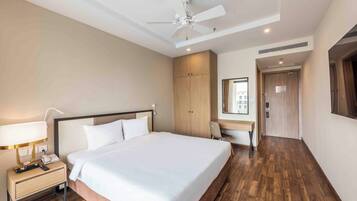Quarto de hotel com cama, mesa de cabeceira, escrivaninha, cadeira, guarda-roupa e ventilador de teto.