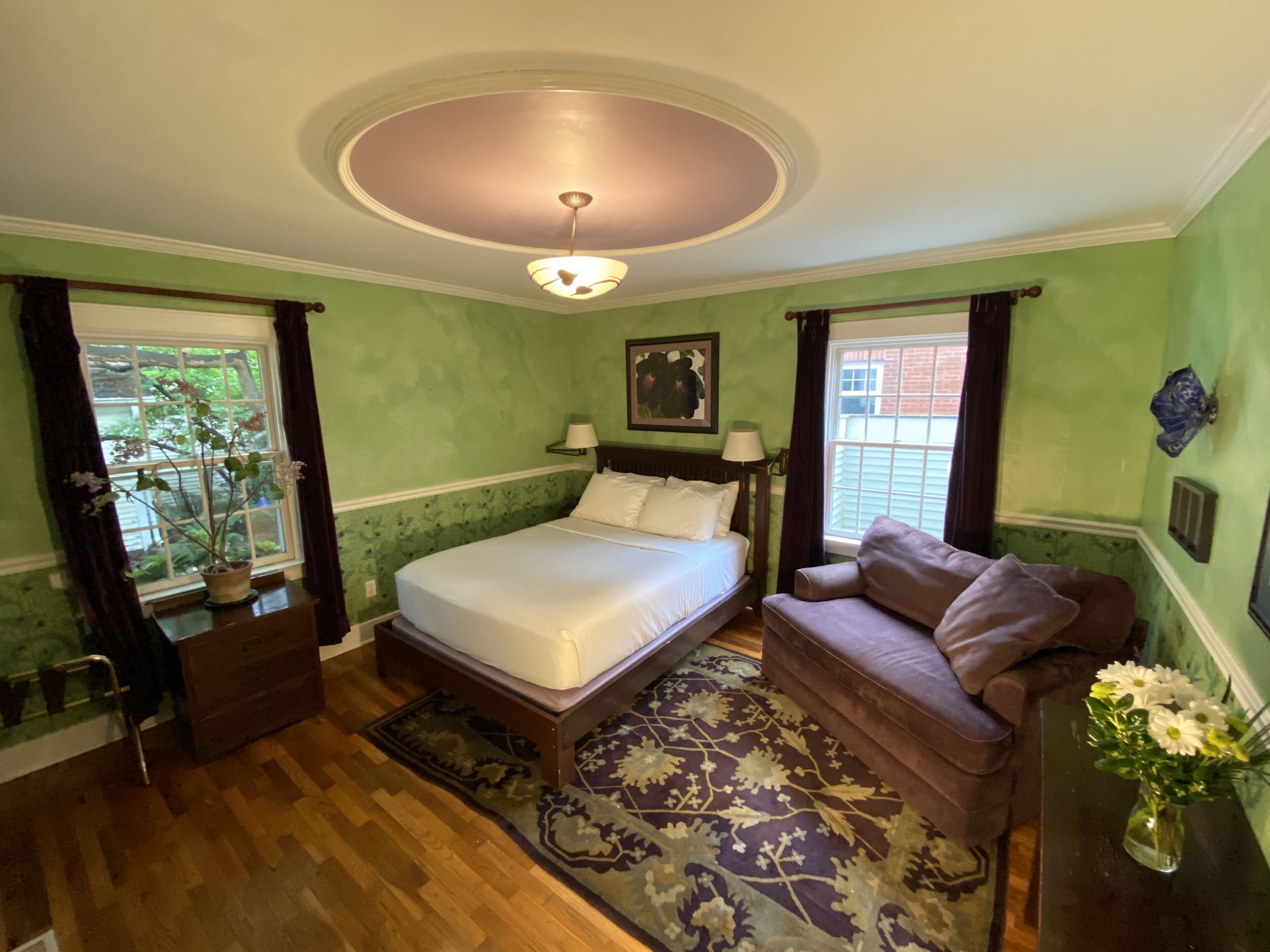 Quarto com cama, mesa de cabeceira, cômoda, janela com cortinas, luminária no teto, um quadro na parede e tapete estampado.