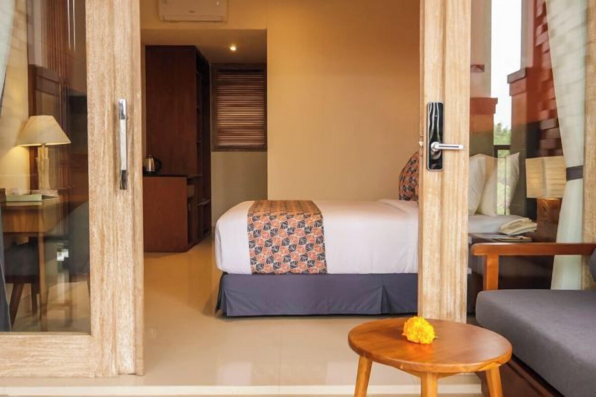 Quarto de hotel com cama, uma mesinha com uma flor, um sofá e uma porta de madeira.