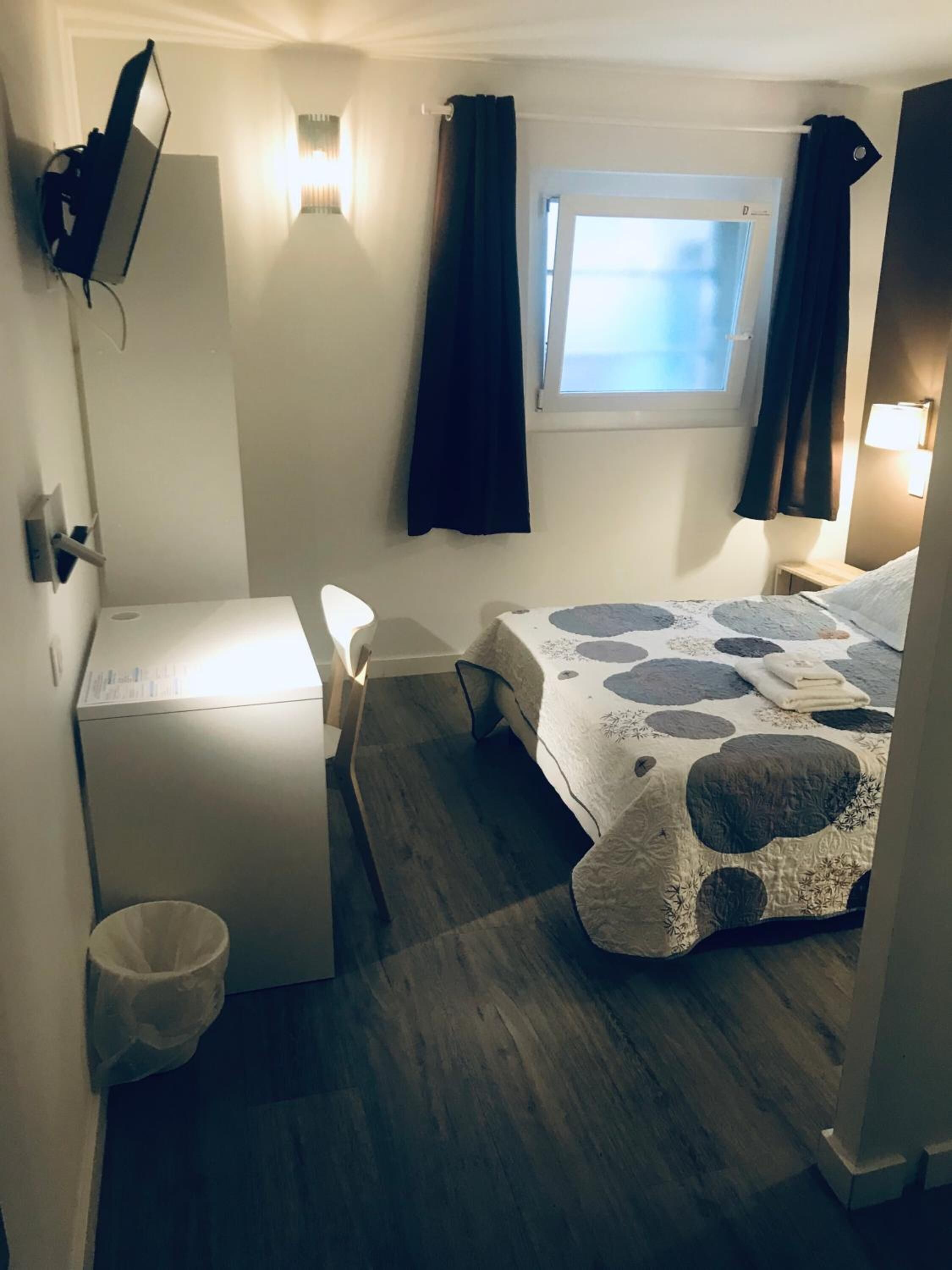 Quarto compacto com cama, escrivaninha pequena, cadeira, máquina de lavar e janela com cortinas.