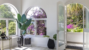 Um quarto claro com janelas amplas, uma planta em vaso e uma pequena mesa com uma escultura.