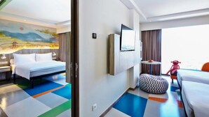 Quarto de hotel com uma cama, uma televisão, uma mesinha com frutas, um banquinho colorido e um mural na parede.