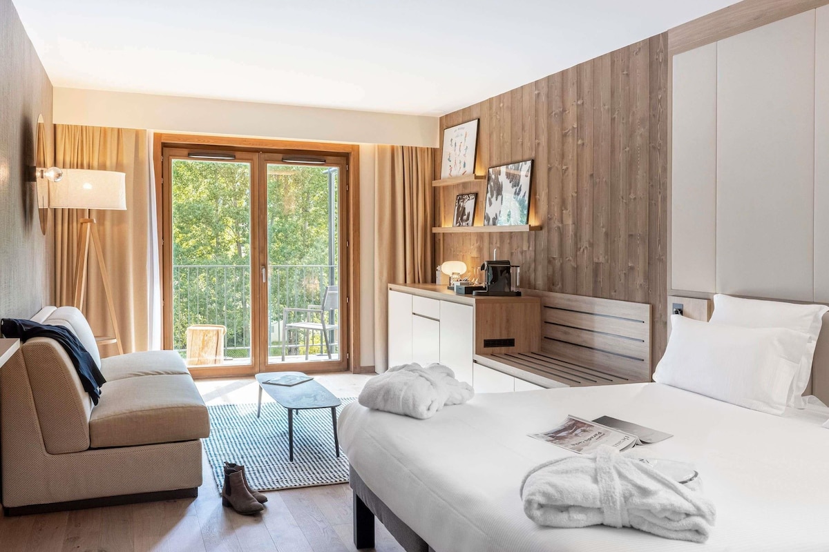 Quarto de hotel com cama, sofá, escrivaninha e varanda com vista para árvores.