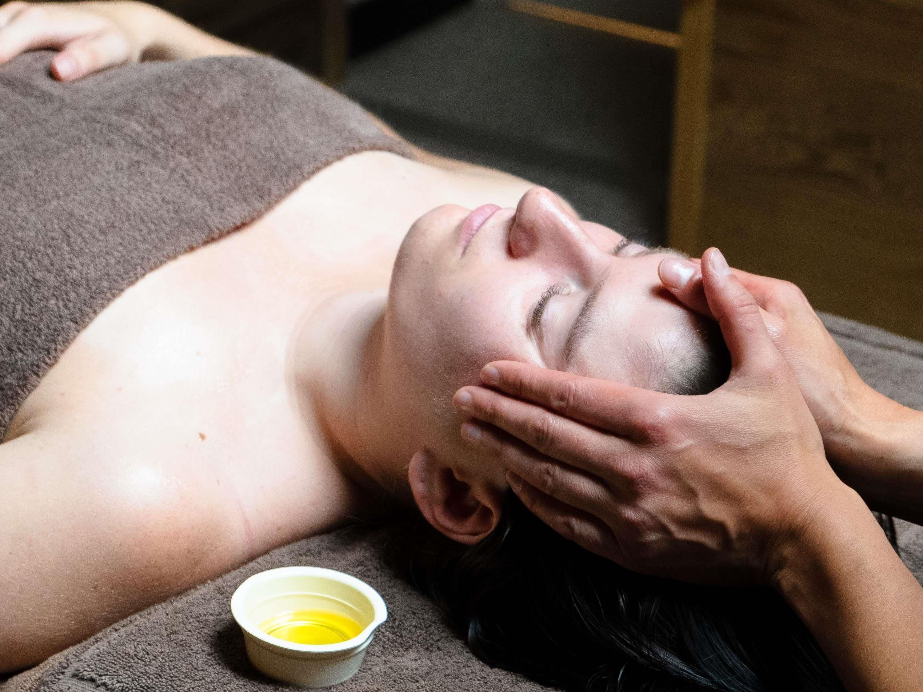 Uma pessoa deitada, com uma toalha cobrindo o tronco, uma tigela com líquido amarelo ao lado e mãos fazendo uma massagem.