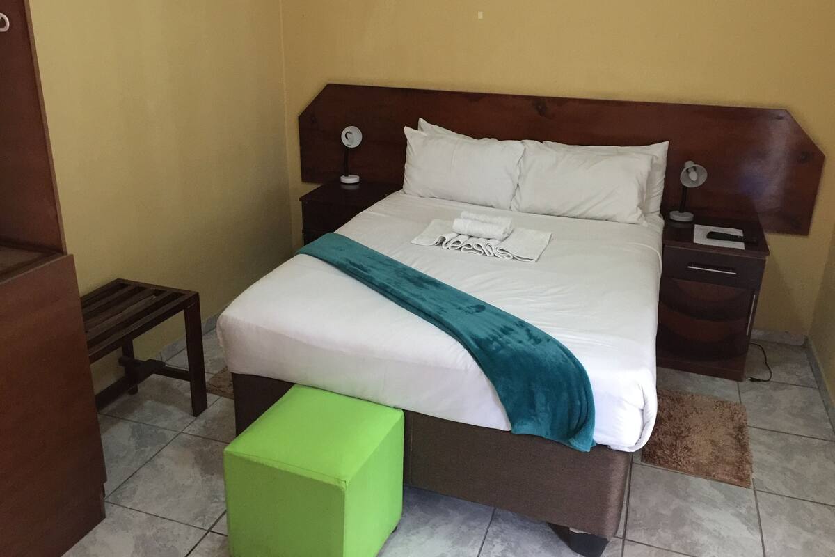Cama arrumada com lençóis brancos, manta azul-esverdeada e toalhas dobradas. Há uma pequena mesa de cabeceira com uma luminária e um criado-mudo com um telefone. Um banquinho verde está posicionado em frente à cama.