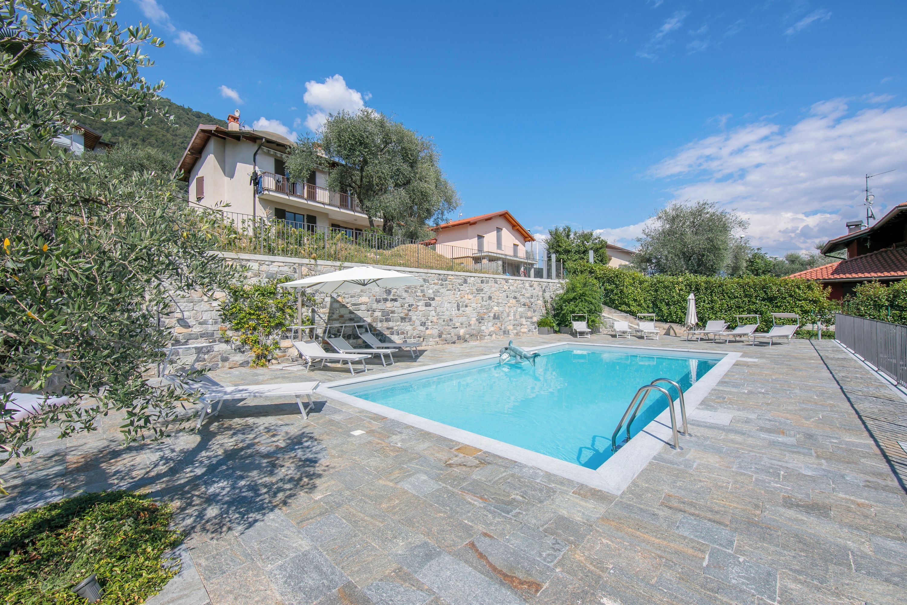 Piscina com espreguiçadeiras, um muro de pedra e vista para casas e árvores.
