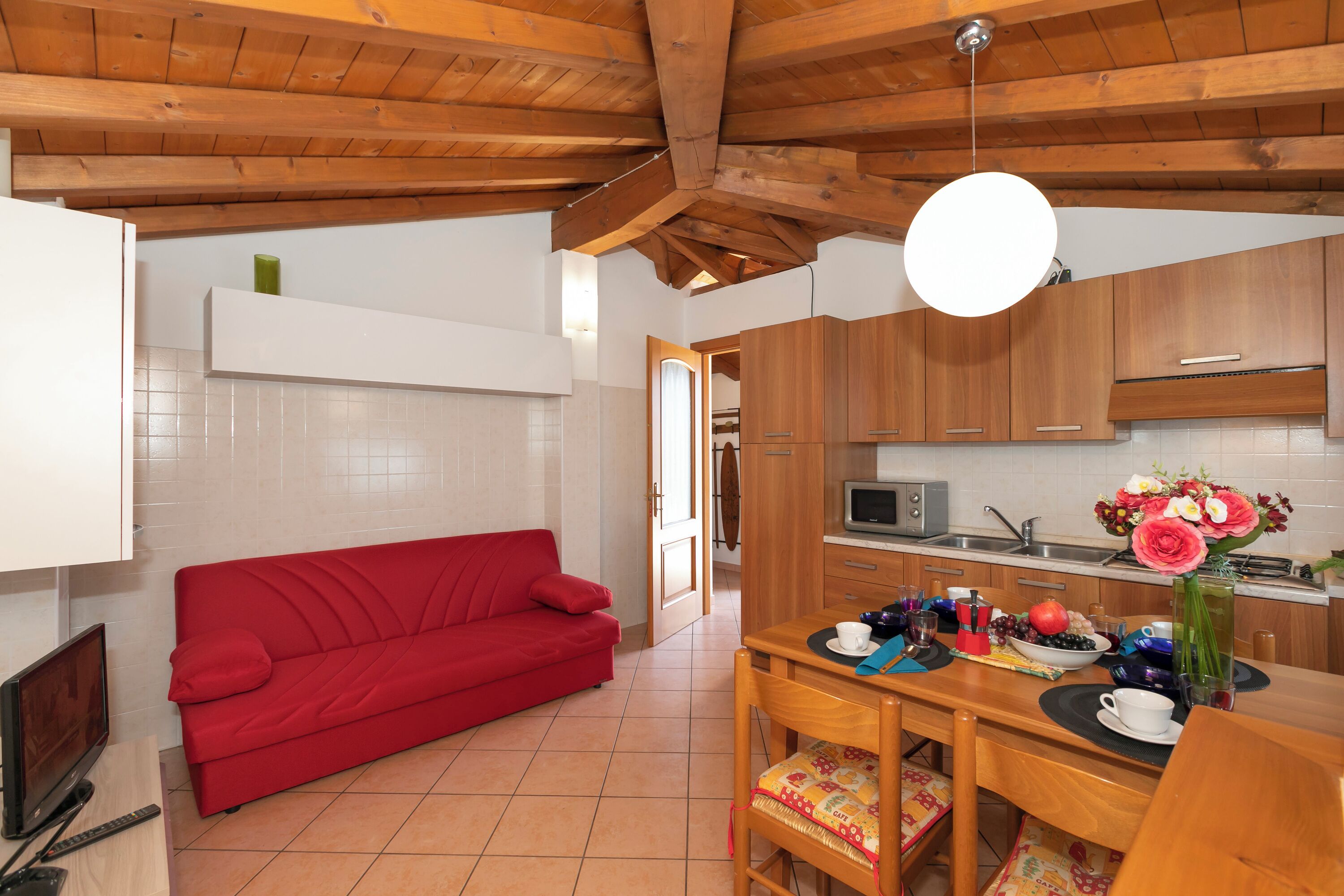 Um quarto aconchegante com um sofá vermelho, vigas de madeira no teto, uma mesa de jantar com um vaso de flores e uma cozinha equipada com micro-ondas e pia.
