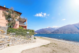 Um edifício costeiro com escadaria de pedra que leva a uma varanda, com vista para um lago e montanhas ao fundo.