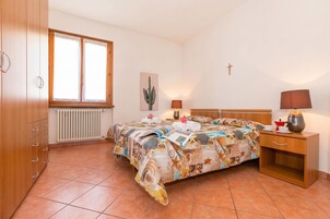 Quarto com cama, mesas de cabeceira, radiador, janela e uma cruz na parede.