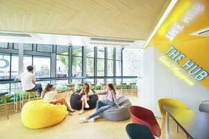 Um interior moderno com amplas janelas, uma parede amarela com a identificação 'THE HUB' e áreas de estar com puffs e cadeiras.