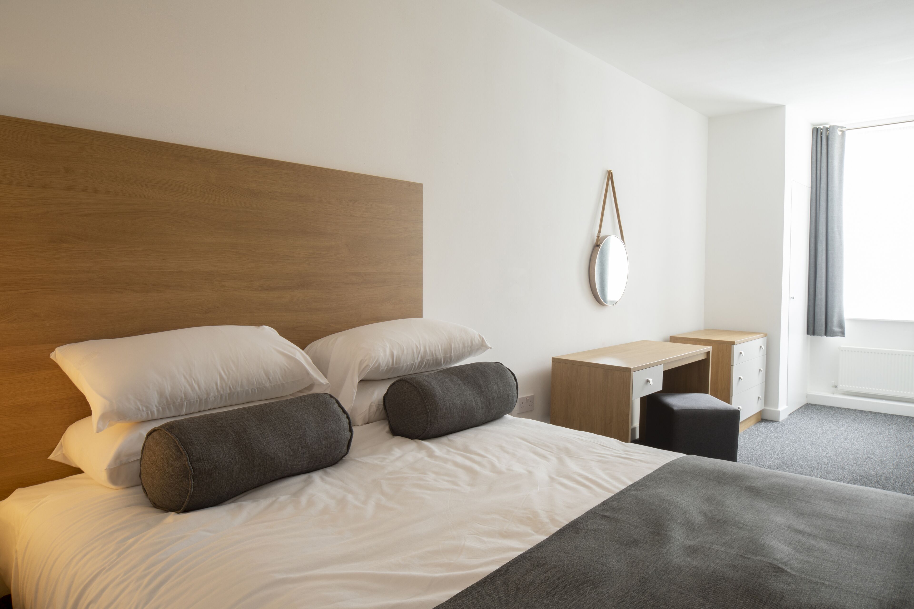 Quarto de hotel moderno com cabeceira de madeira, cama com roupa de cama branca e cinza, uma pequena escrivaninha e um espelho na parede.