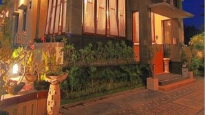 Uma casa de dois andares com uma janela grande na fachada, um pequeno alpendre e um jardim com plantas em vasos.