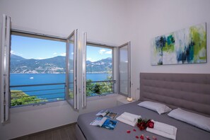 Um quarto com uma cama, janela com vista para um lago e montanhas, e um quadro na parede.