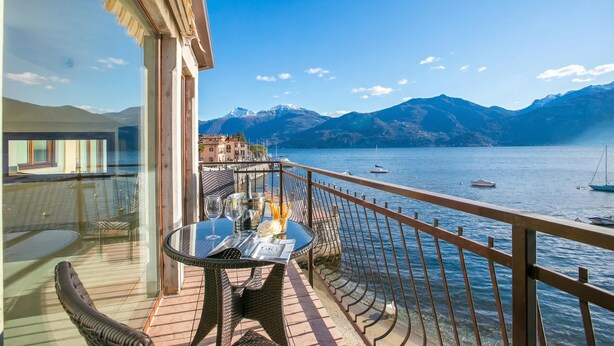 Uma varanda com mesa e cadeiras, vista para um lago com barcos e montanhas ao fundo.