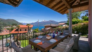 Mesa de jantar montada ao ar livre, com cadeiras de vime, vista para montanhas e um lago.
