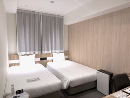 Quarto de hotel com duas camas, uma escrivaninha, uma cadeira, uma mesinha, um abajur e uma luminária no teto.