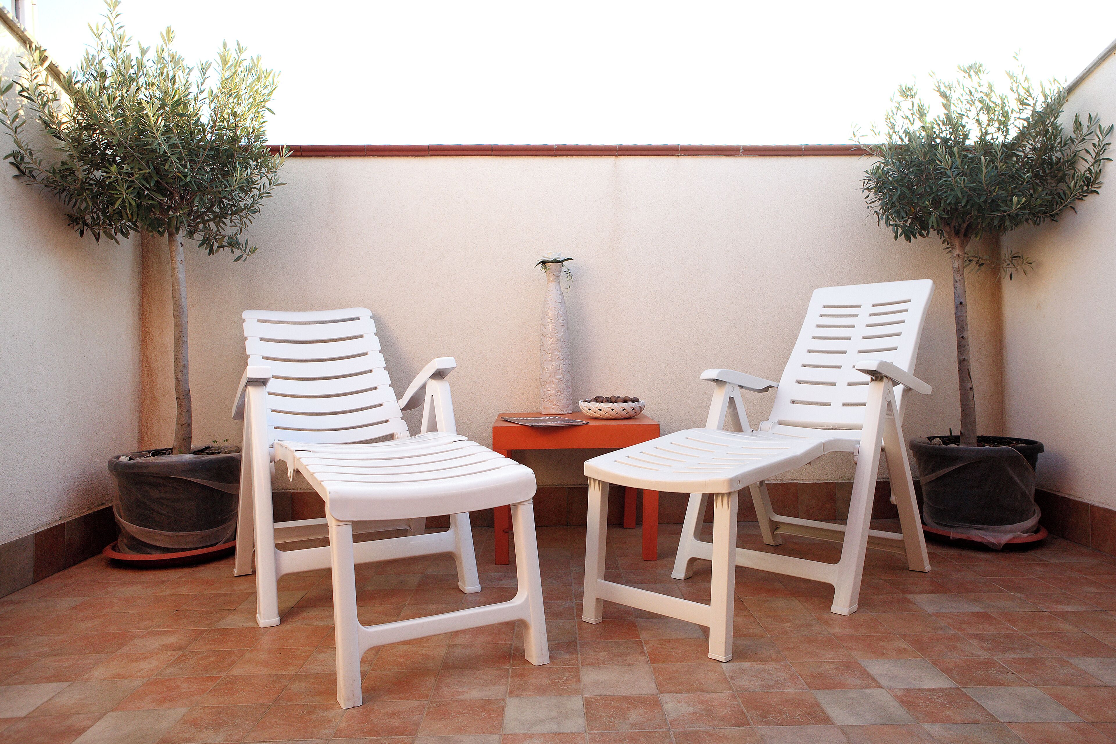 Duas cadeiras de plástico branco e uma pequena mesa em área externa com piso de azulejos e plantas em vasos.