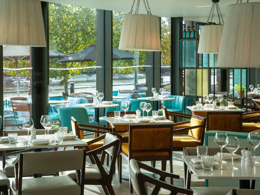 Um restaurante com mesas, cadeiras, janelas amplas e luminárias pendentes.