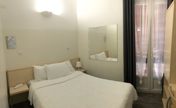 Quarto de hotel com uma cama, mesa de cabeceira com luminária, espelho e janela com persianas.