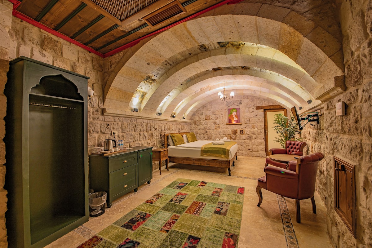 Quarto com parede de pedra, uma cama, guarda-roupa verde, mesa de cabeceira de madeira e uma poltrona vermelha.