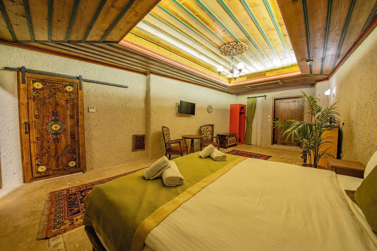 Quarto de hotel com teto de madeira, cama com colcha verde, uma pequena mesa com cadeiras, televisão e uma planta.