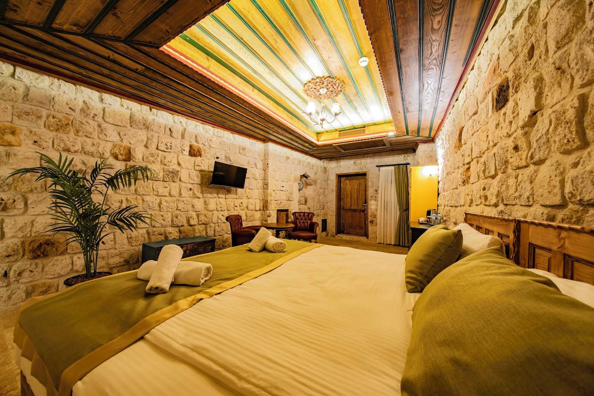 Quarto com parede de pedra, vigas de madeira no teto, cama com roupa de cama verde, planta em vaso e televisão na parede.