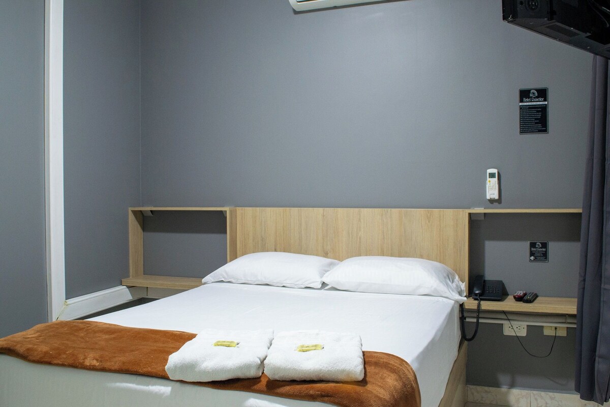 Quarto de hotel com cama, mesa de cabeceira, telefone e ar condicionado fixado na parede.