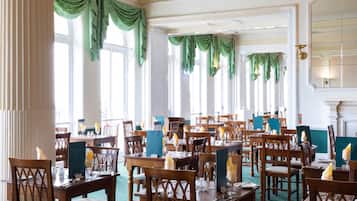 Uma sala de jantar com várias mesas e cadeiras de madeira, cortinas verdes e um candelabro.
