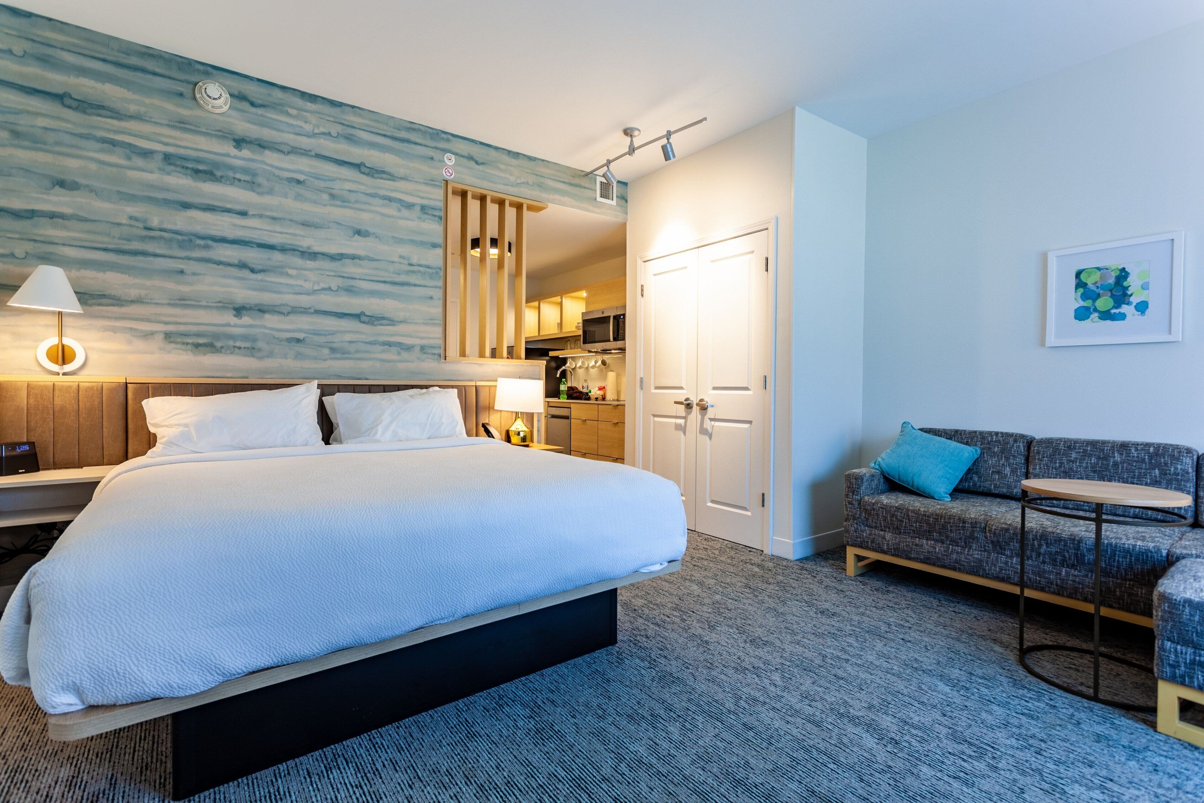 Quarto de hotel com uma cama grande, um sofá, uma mesinha e uma cozinha compacta ao fundo.