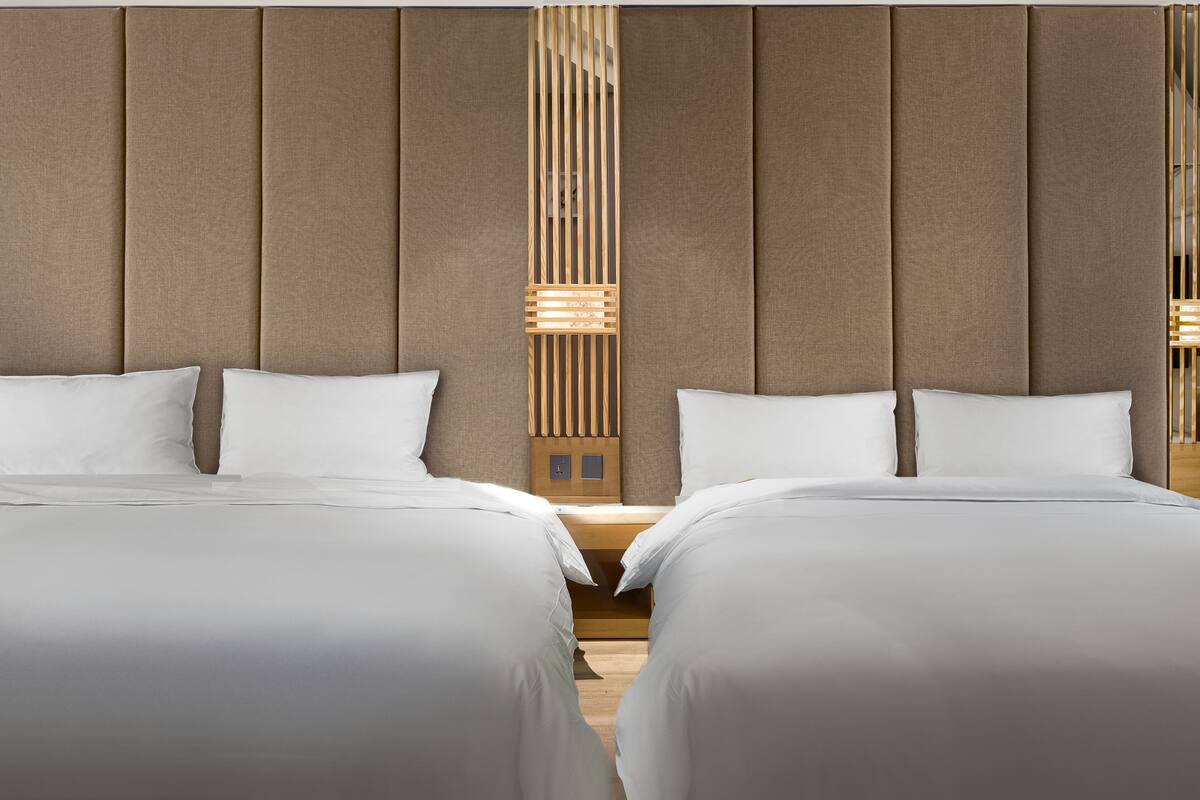 Quarto de hotel com duas camas, cabeceira de madeira e mesa de cabeceira com abajur.