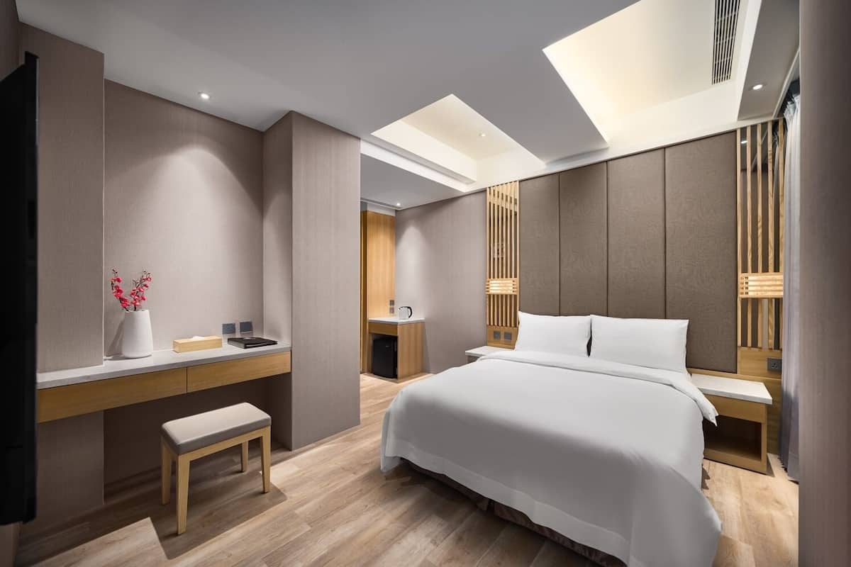 Quarto de hotel moderno com uma cama grande, uma mesinha de cabeceira, um banco e uma escrivaninha com um vaso de flores.