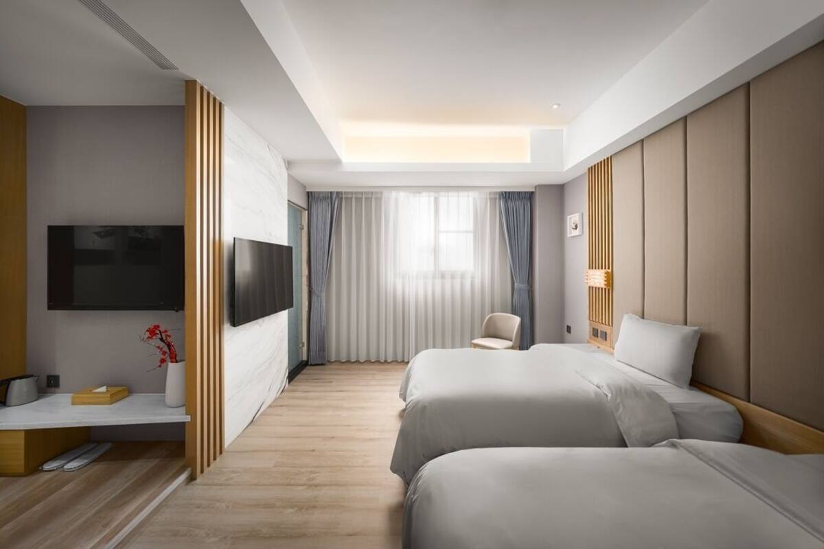 Quarto de hotel com duas camas, televisão, escrivaninha com cadeira e janela com cortinas.