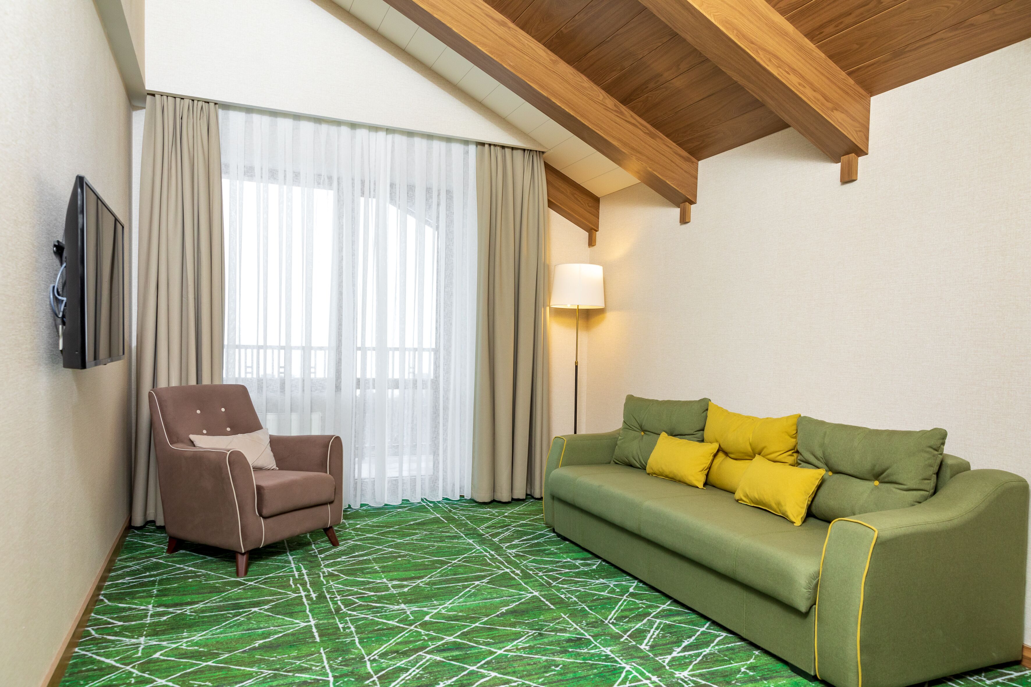 Um quarto com carpete verde estampado, uma poltrona marrom, um sofá verde com almofadas amarelas, uma luminária e uma televisão fixada na parede.