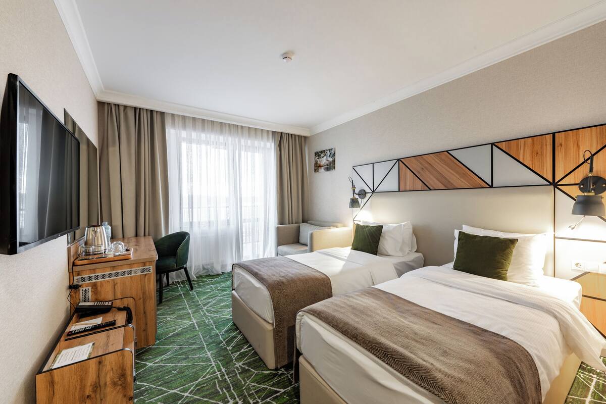 Quarto de hotel com duas camas, uma escrivaninha, uma cadeira, televisão de tela plana e uma janela grande com cortinas.