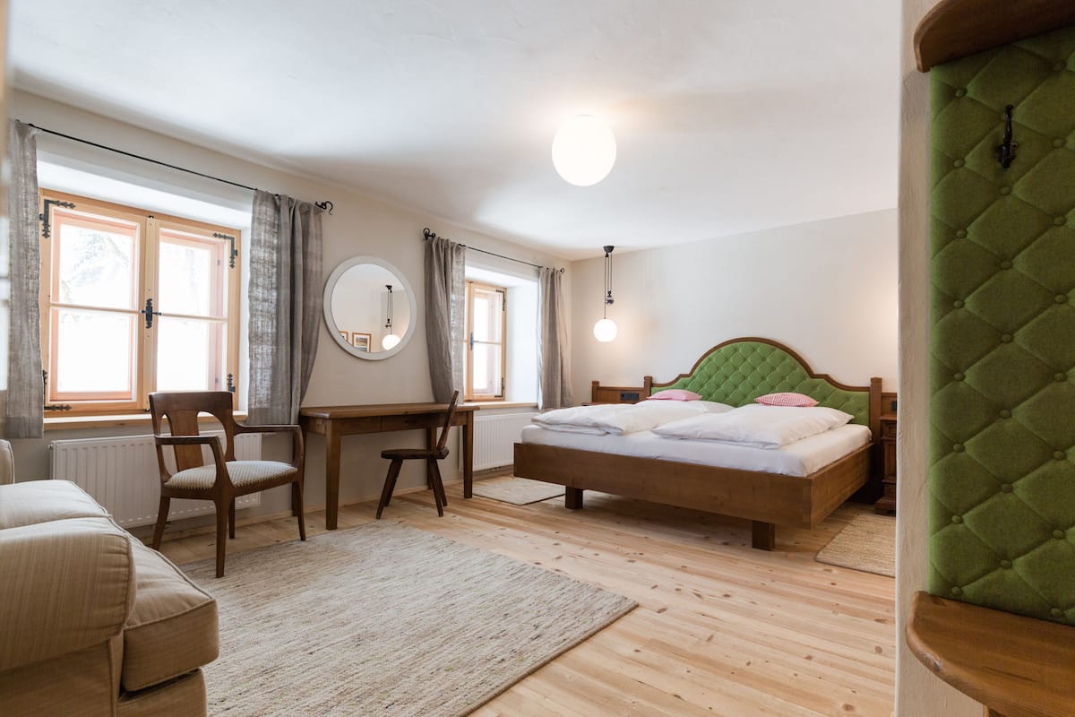 Quarto com cama de madeira, escrivaninha, cadeira, espelho redondo e janela com cortinas.