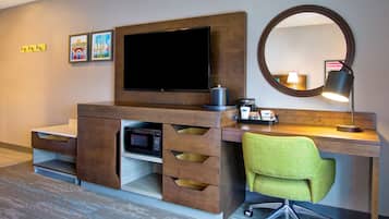 Quarto de hotel com uma secretária de madeira, uma cadeira verde, uma televisão de tela plana, uma máquina de café e um espelho redondo.