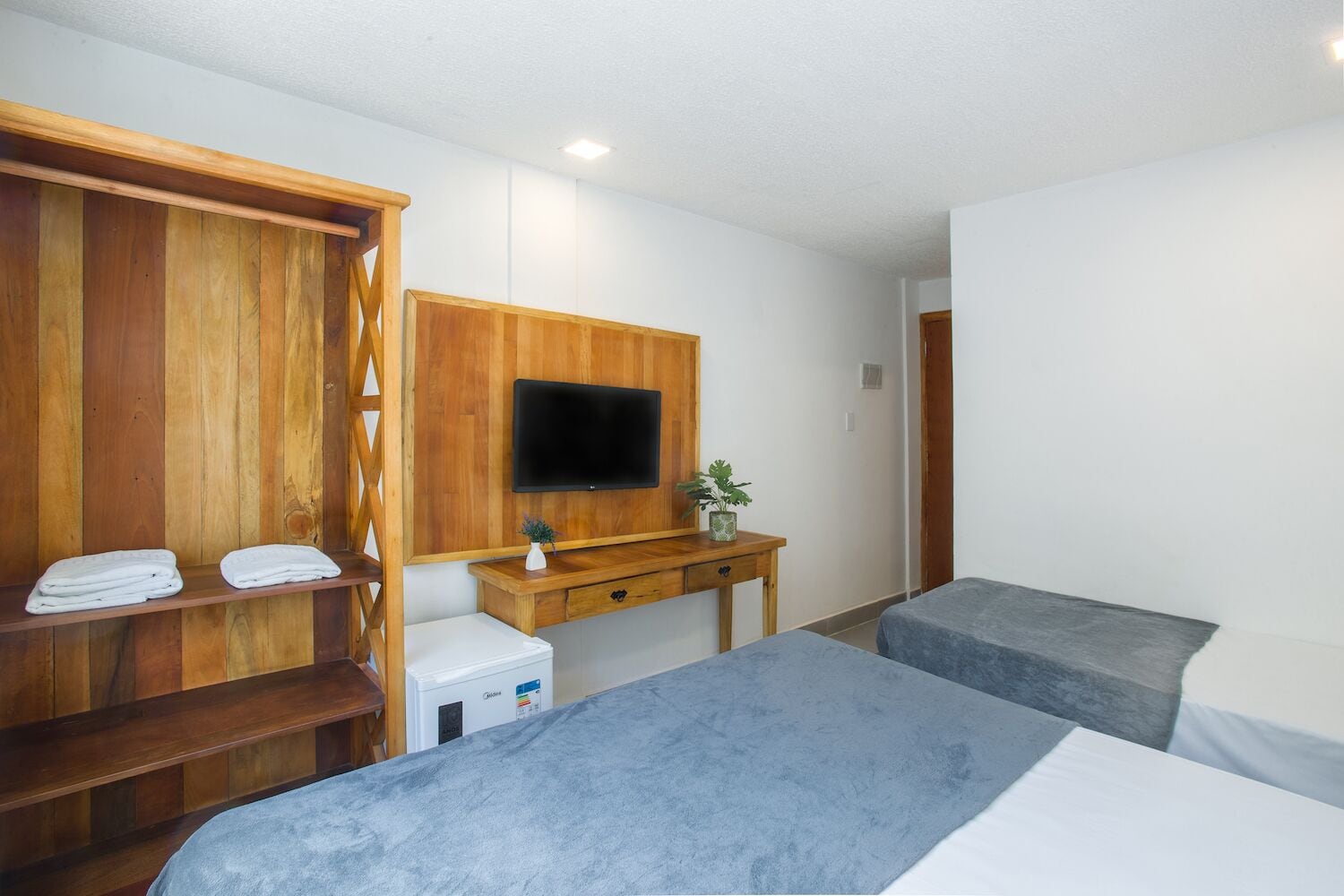 Quarto de hotel com guarda-roupa de madeira, televisão de ecrã plano fixada num painel de madeira, secretária com uma planta e duas camas com roupa de cama cinzenta.
